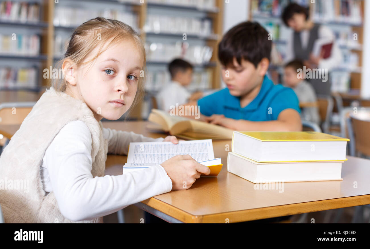 Intelligent tween girl reading in school library on background with ...