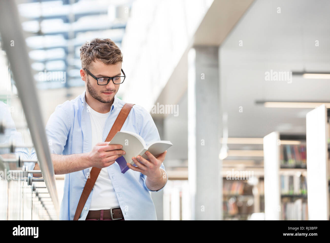 Young student reading book while standing by railing at corridor in ...