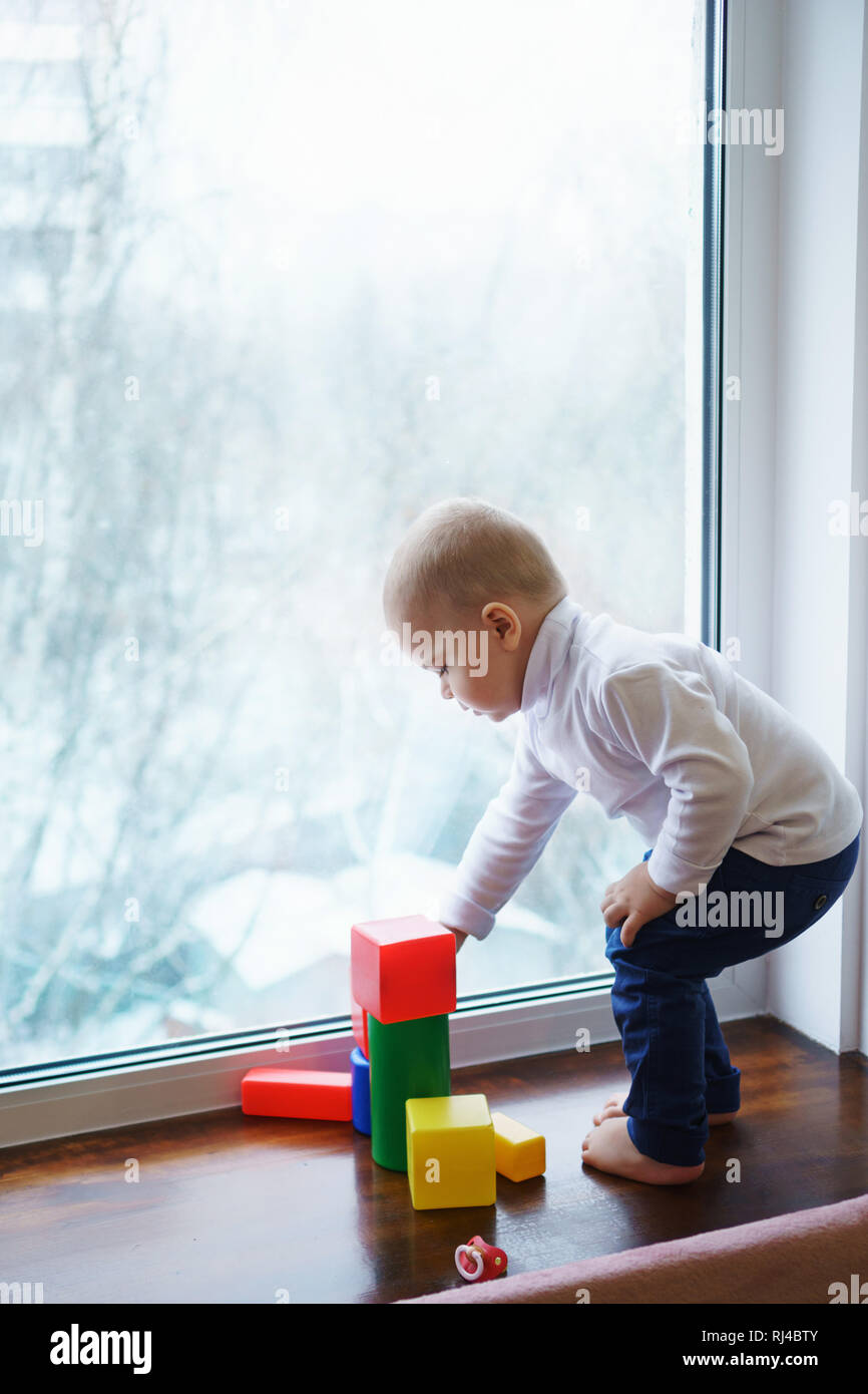 Little boy stands barefoot on windowsill. He plays with colored cubes ...