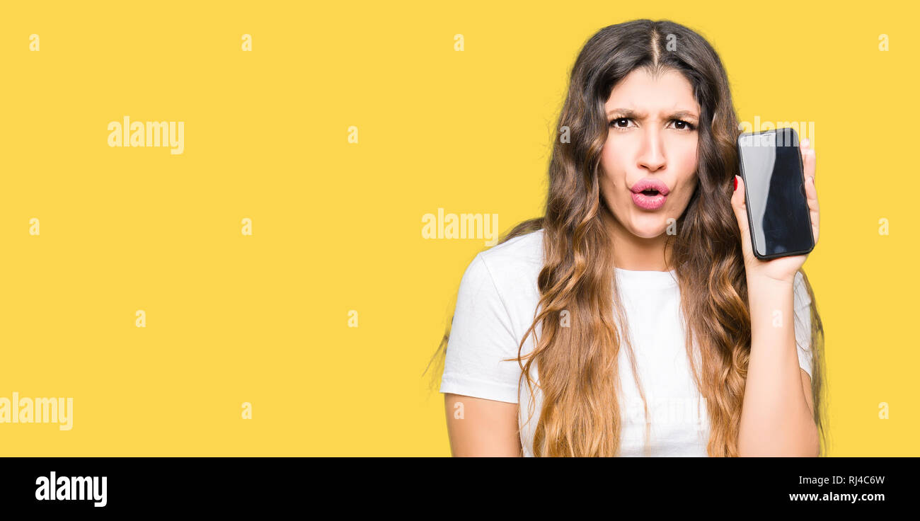 Young adult woman showing smartphone screen scared in shock with a ...