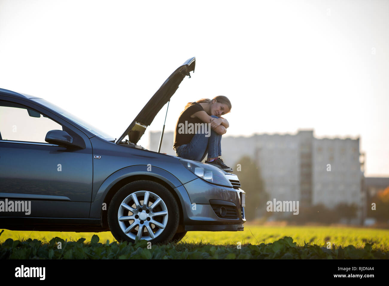 Young attractive sad woman sitting on car with popped hood in green ...