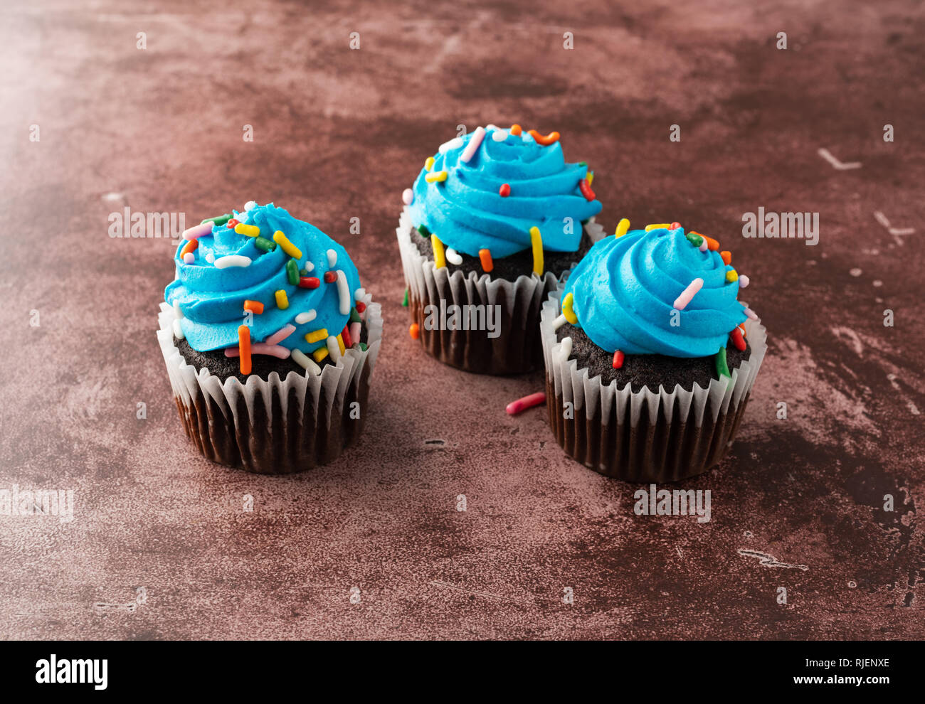 Side view of three blue frosted chocolate cupcakes on a maroon ...