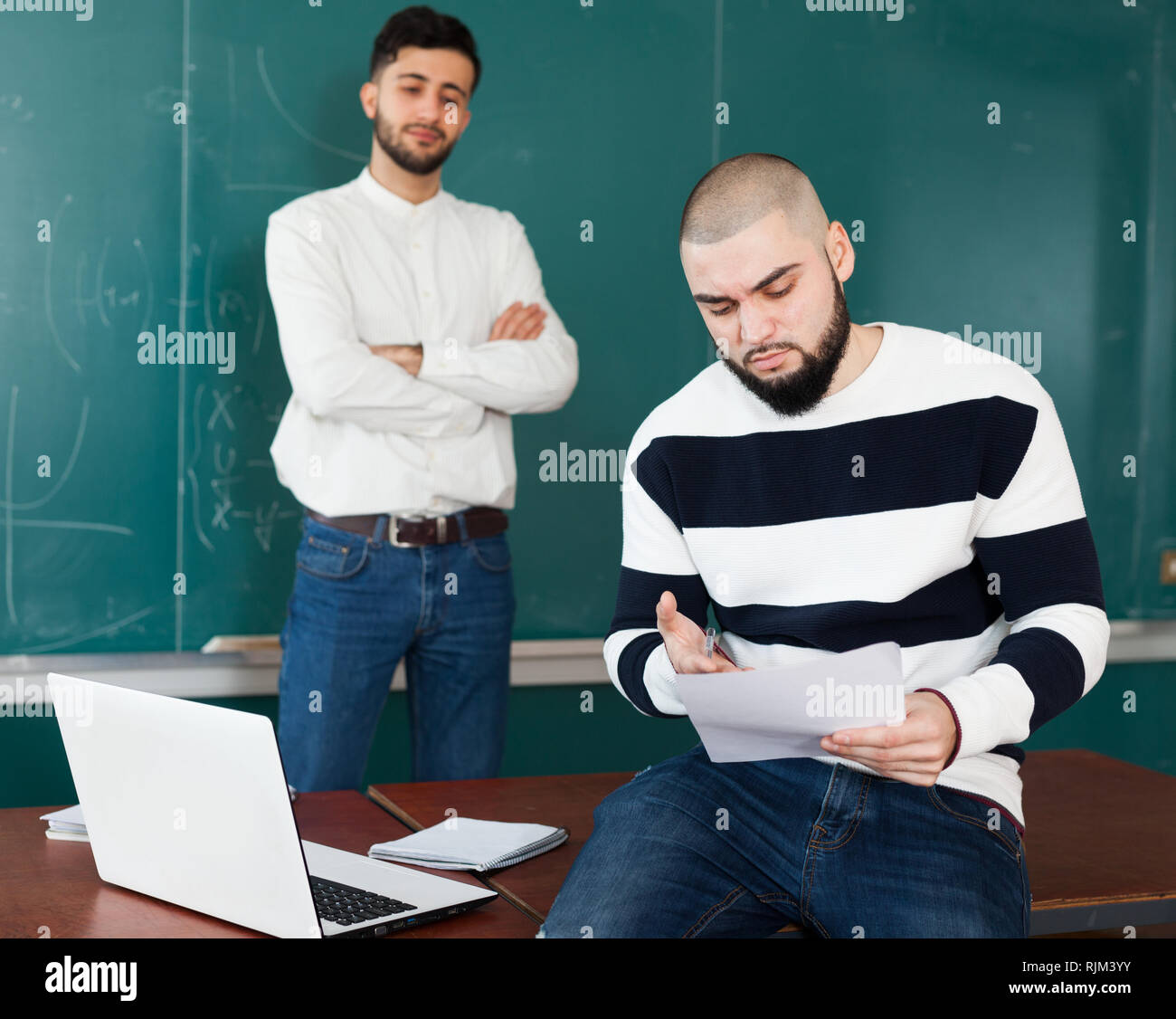 Two young handsome students working with laptop near blackboard at ...