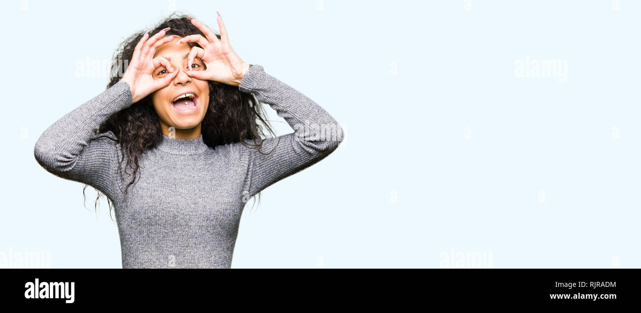 Young beautiful girl with curly hair doing ok gesture like binoculars ...