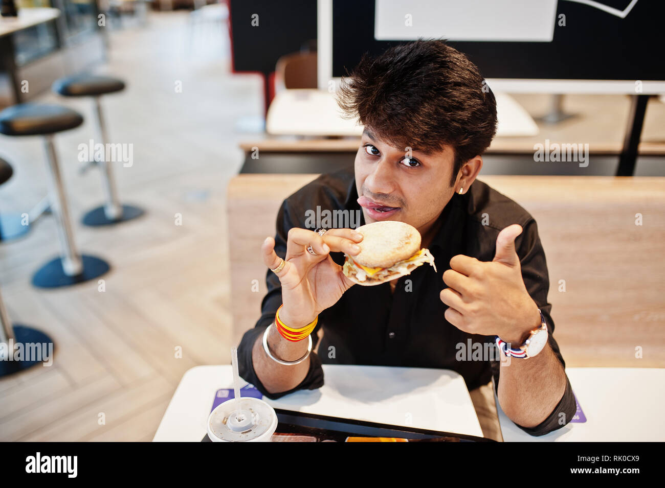 Stylish indian man sitting at fast food cafe and eating hamburger. Show ...