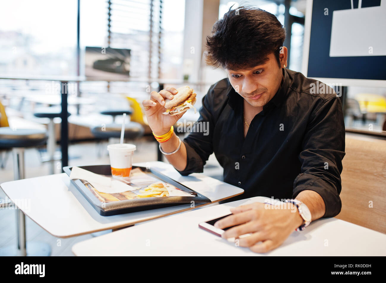 Stylish indian man sitting at fast food cafe and eating hamburger, read ...