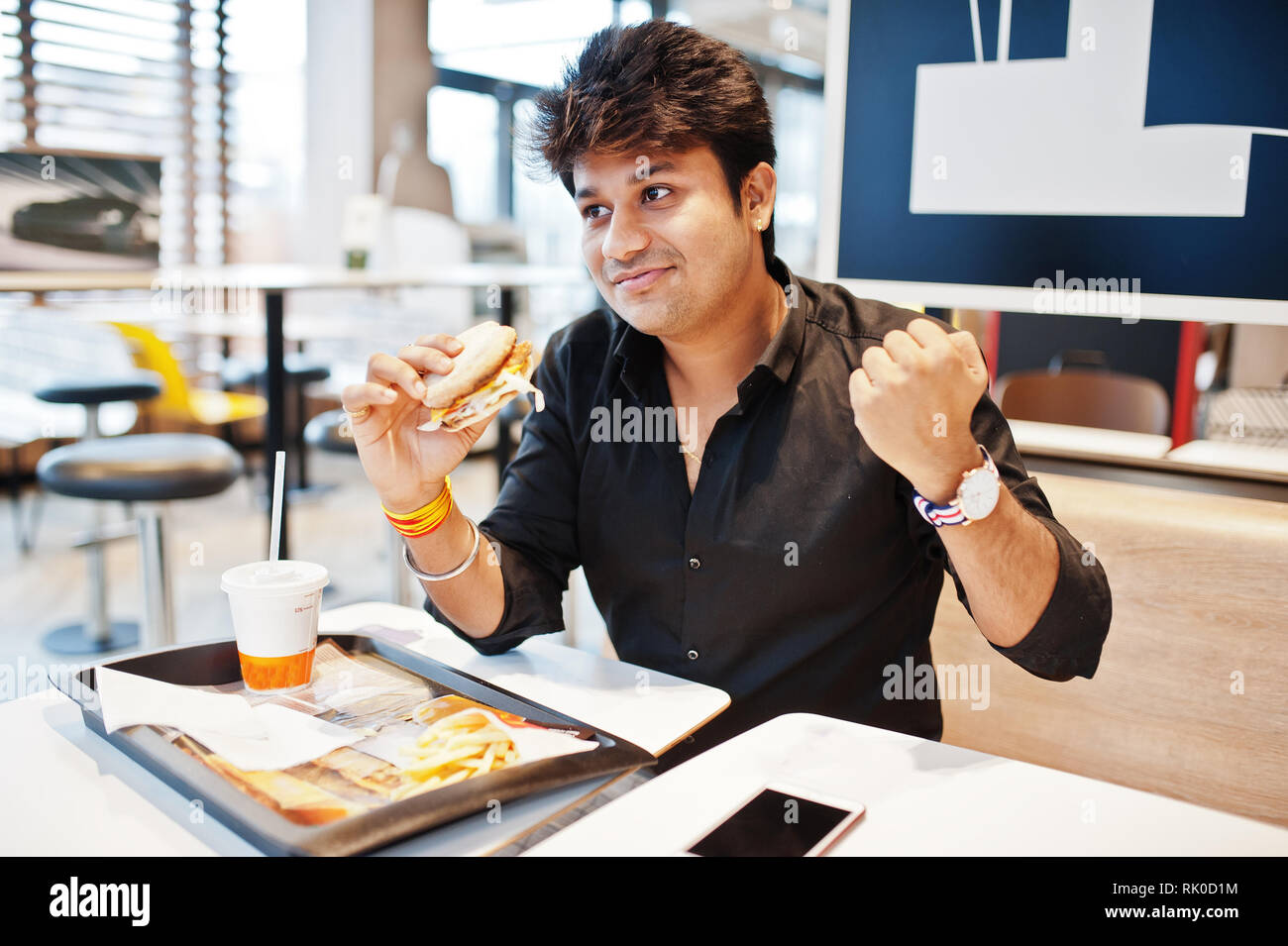 Stylish indian man sitting at fast food cafe and eating hamburger, read ...