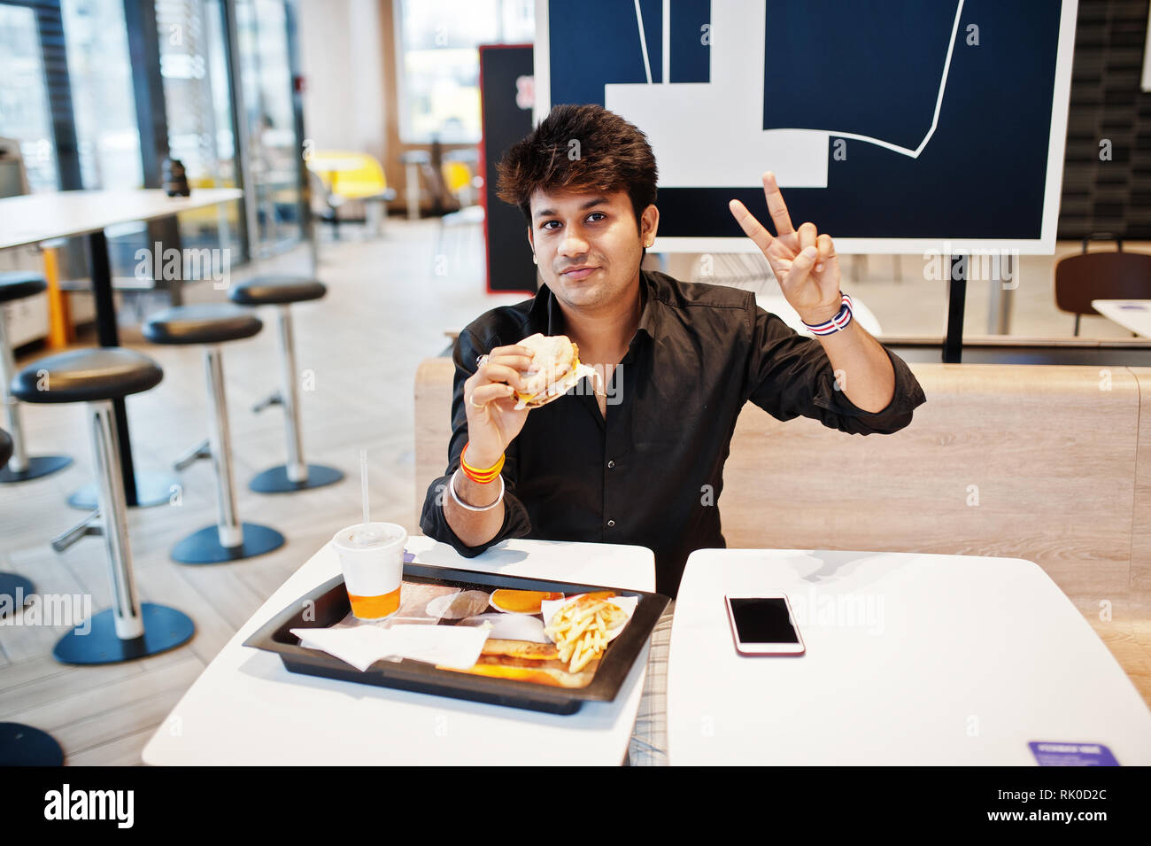 Stylish indian man sitting at fast food cafe and eating hamburger and ...