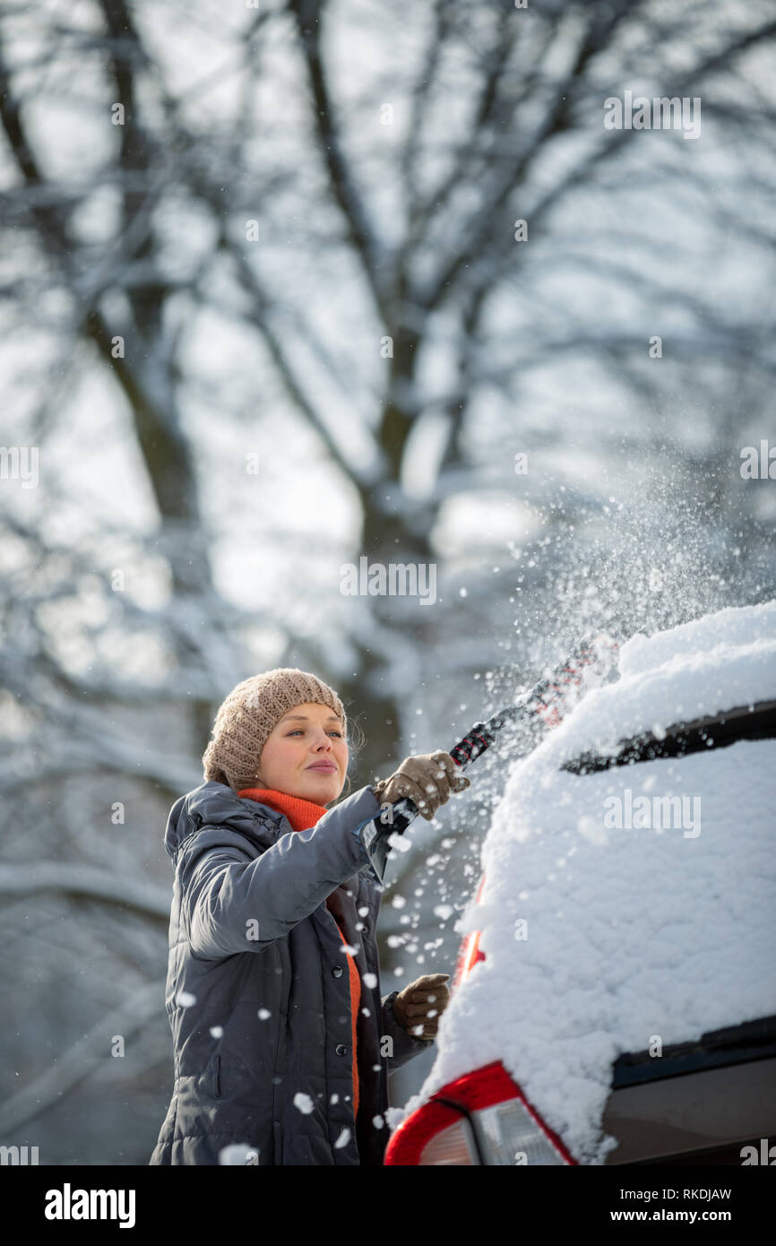 Pretty, young woman cleaning her car from snow after heavy snowstorm ...