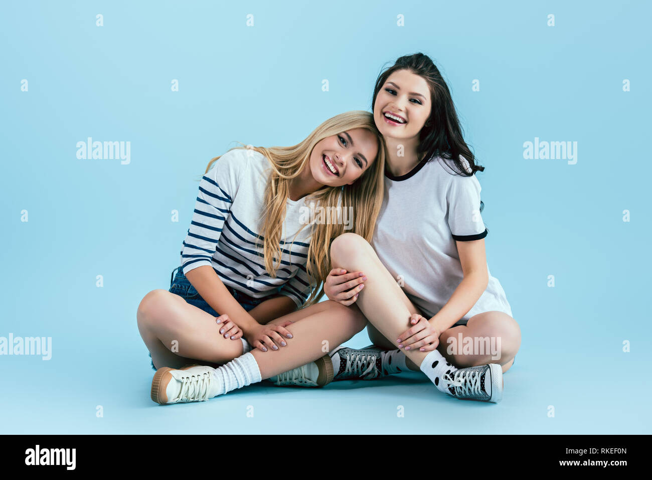 Laughing girls sitting on floor with legs folded on blue background ...