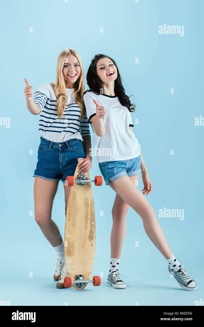Stylish girls with wooden longboard posing with thumbs up on blue ...