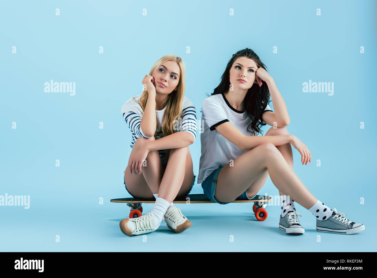 Pensive girls sitting on longboard and propped faces with hands on blue ...