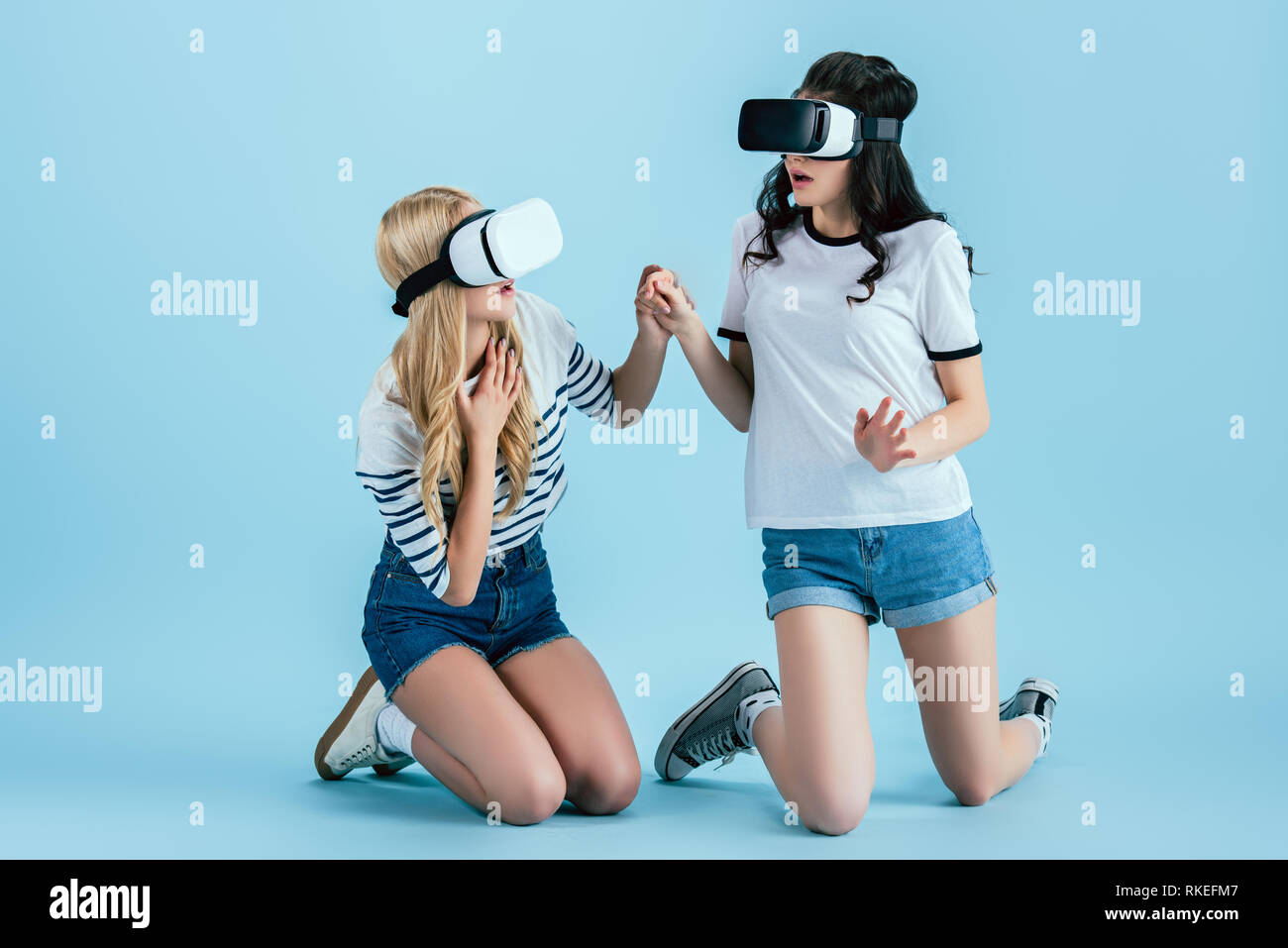 Scared girls in VR headset standing on knees and holding hands on blue ...