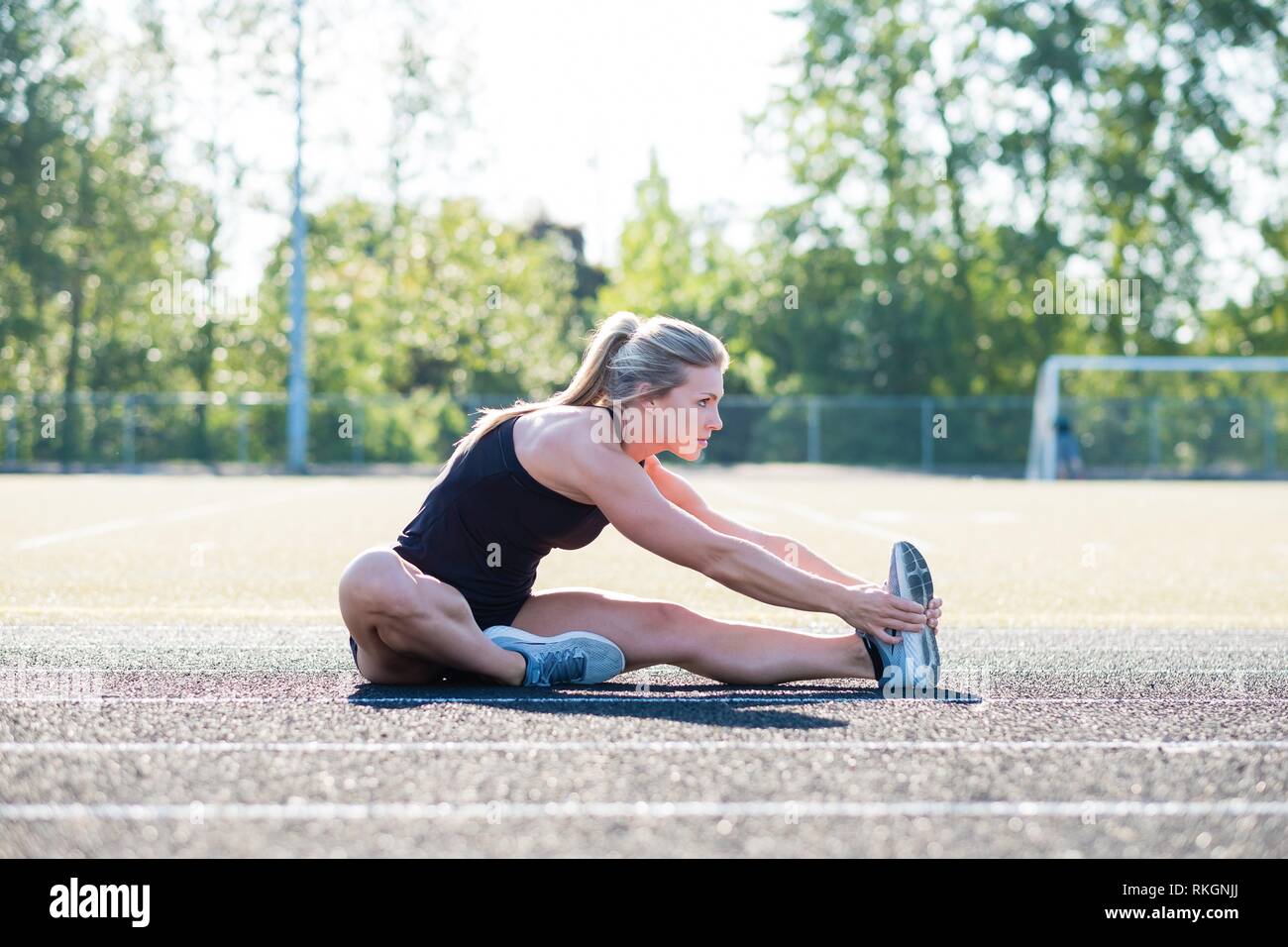 Female runner stretches sitting down on an outdoor track in the summer ...