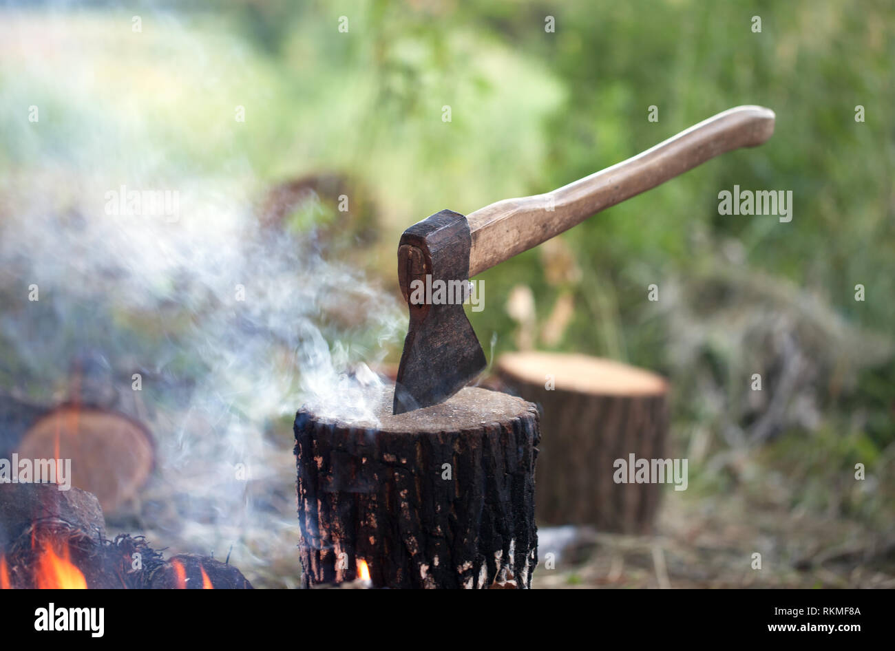 Axe in tree stump and campfire with smoke in summer forest at sunny day ...