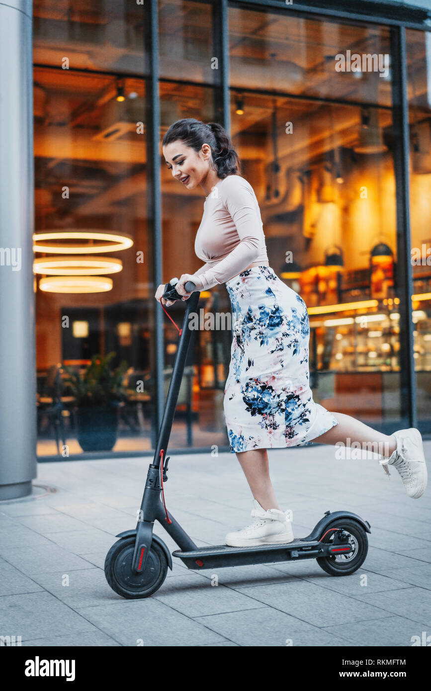 Attractive young woman riding a kick scooter at cityscape background ...