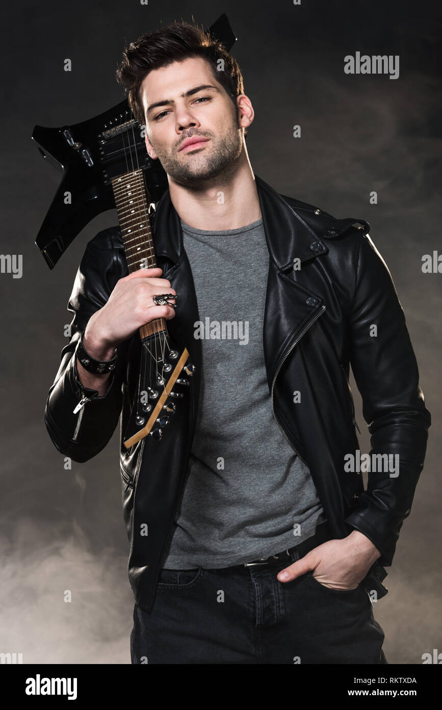 handsome rocker holding electric guitar and looking at camera on dark ...