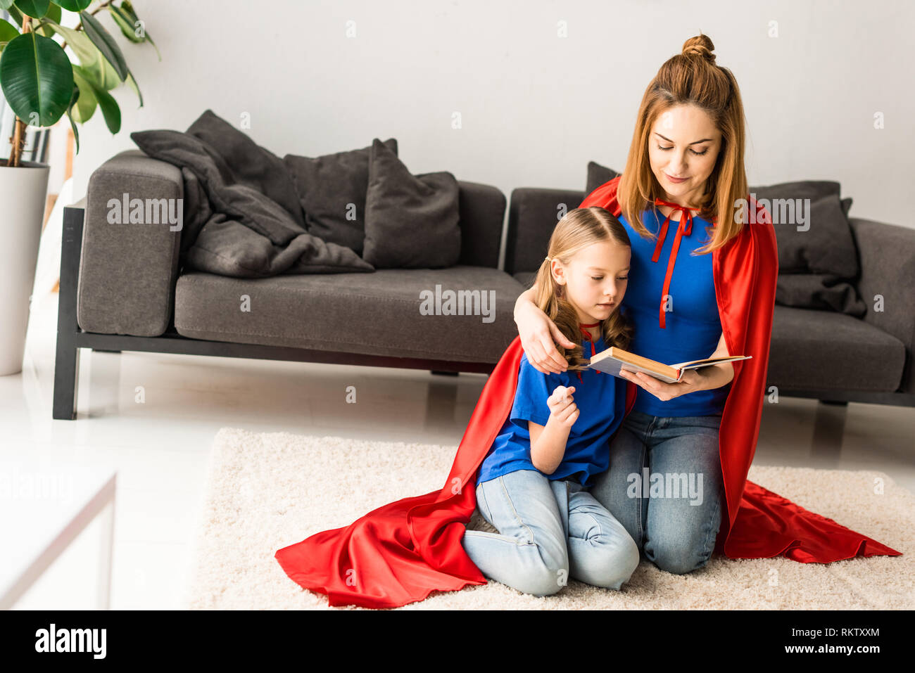 Cute kid and mother in red cloaks sitting on floor and reading book ...