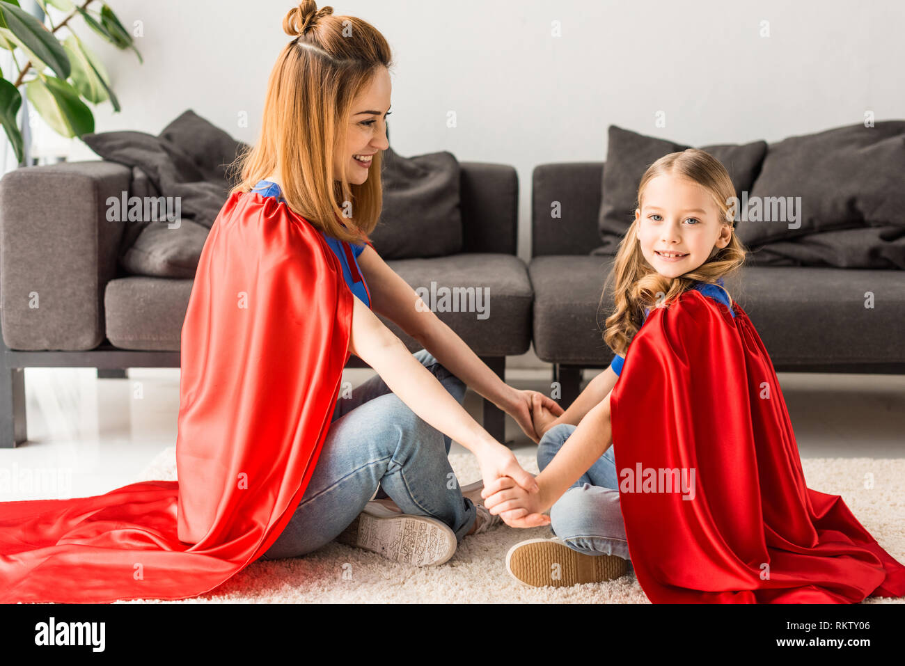 Cute kid and mother in red cloaks sitting on floor and playing at home ...