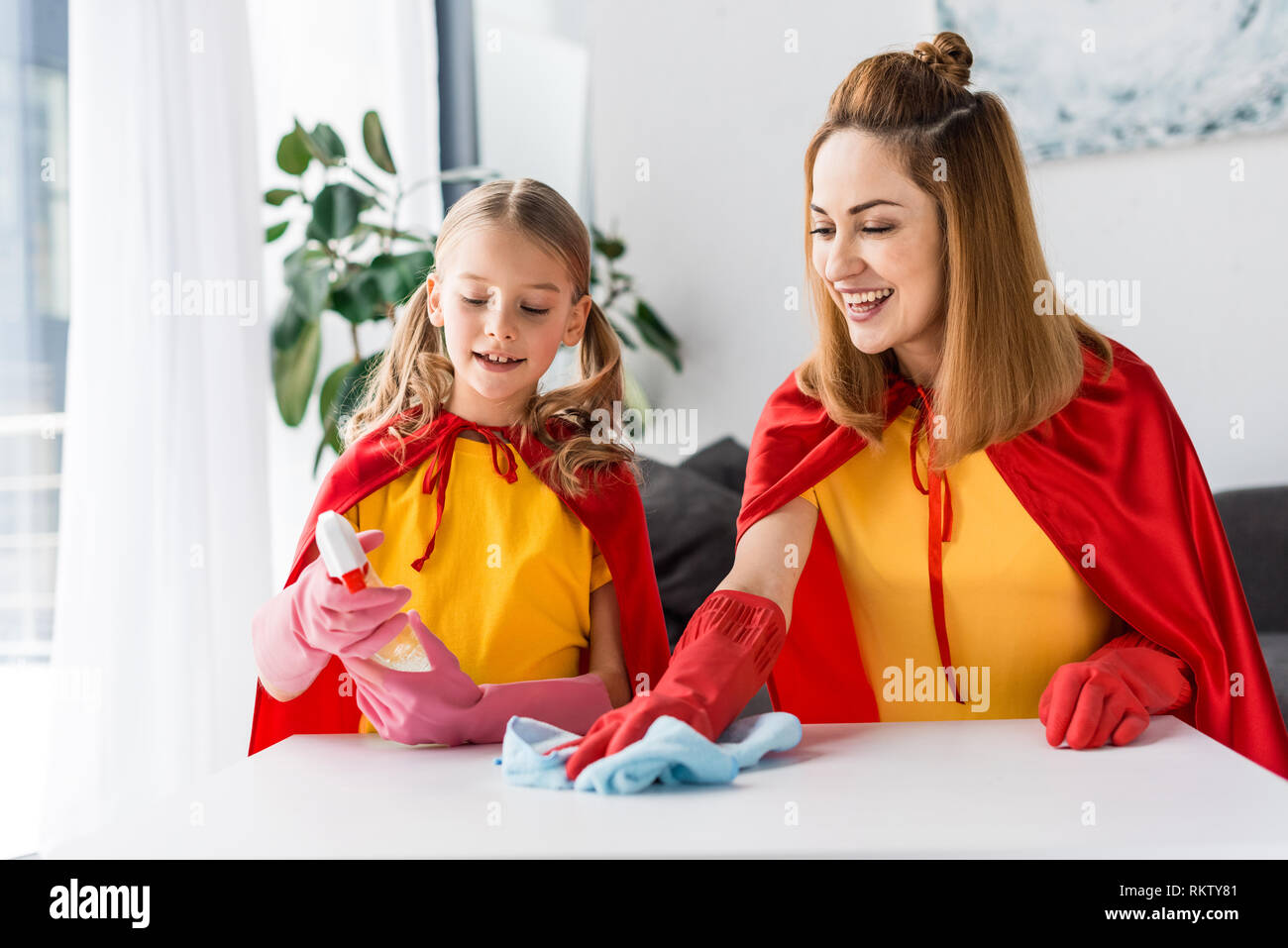 Mother and daughter in red capes and rubber gloves dusting at home ...