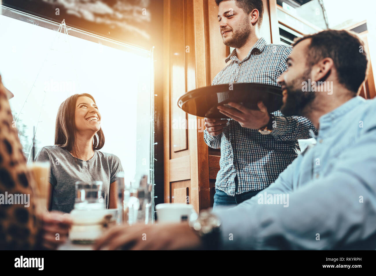 A young handsome waiter is serving a group of young smiling friends in ...