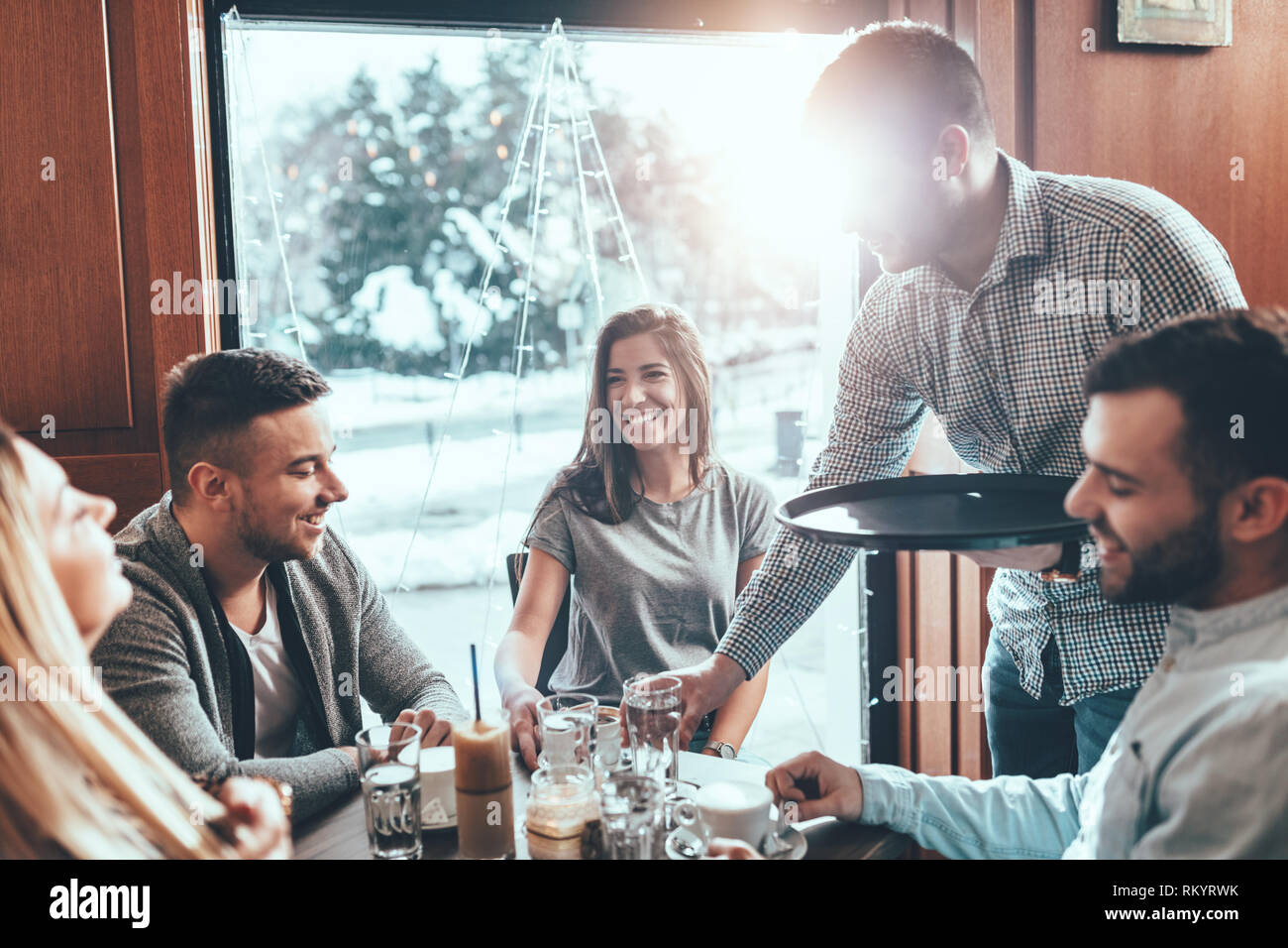 A young handsome waiter is serving a group of young smiling friends in ...