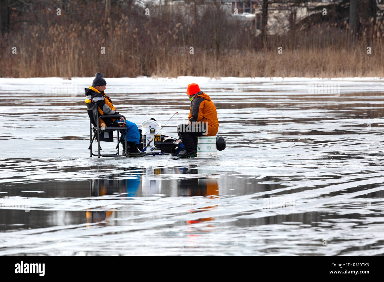 Two men ice fishing on the lake while sitting on a bucket and chair ...