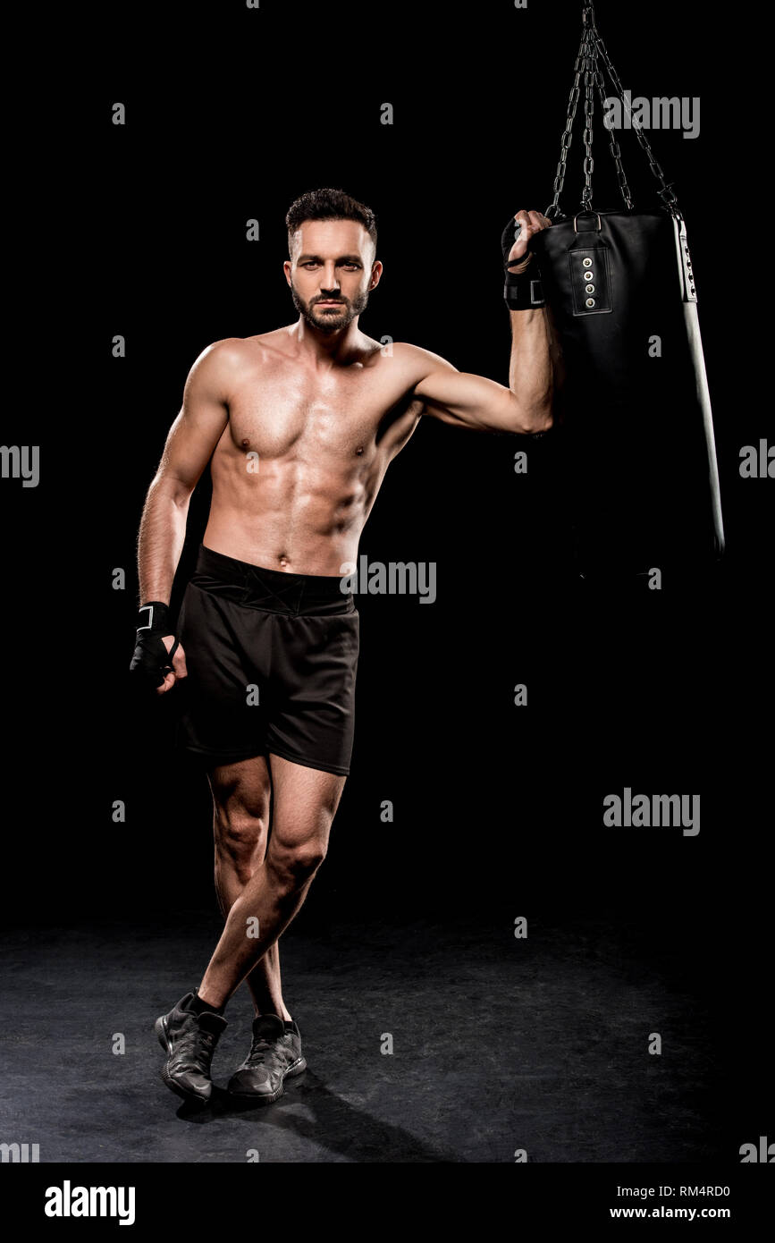 handsome boxer standing with crossed legs near punching bag on black ...