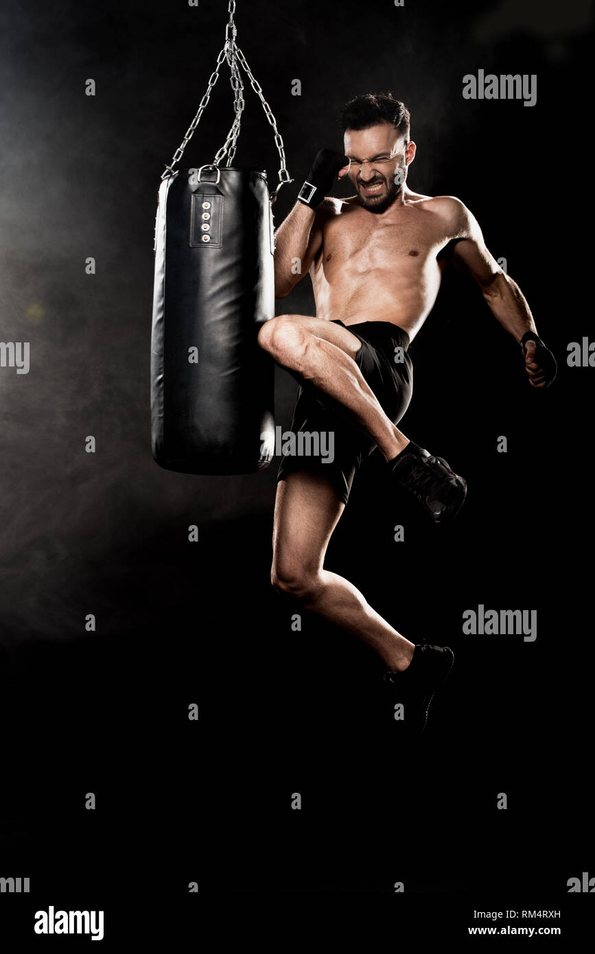 athletic boxer performing flying kick near punching bag on black with ...