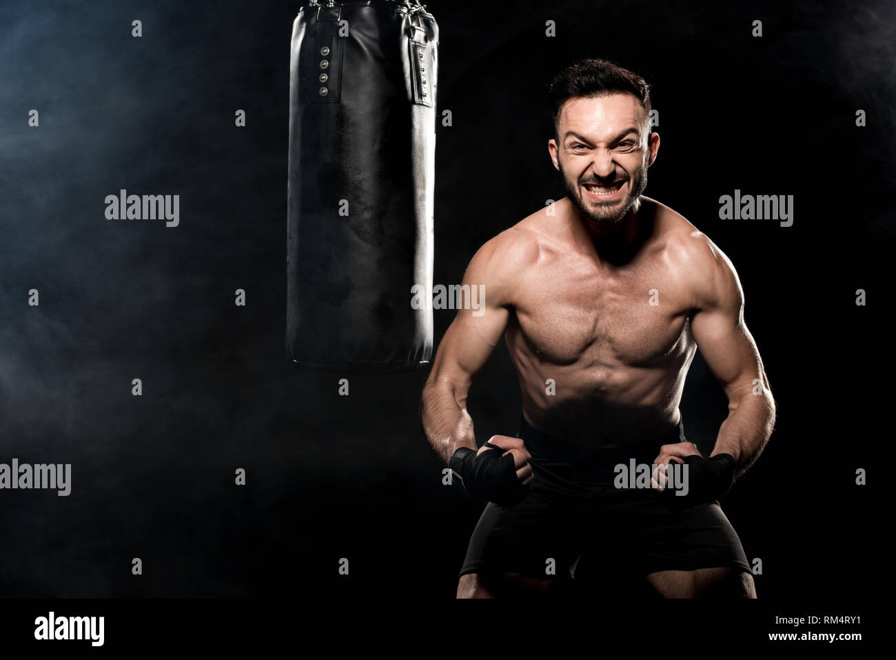 angry boxer showing muscles punching bag on black with smoke Stock ...