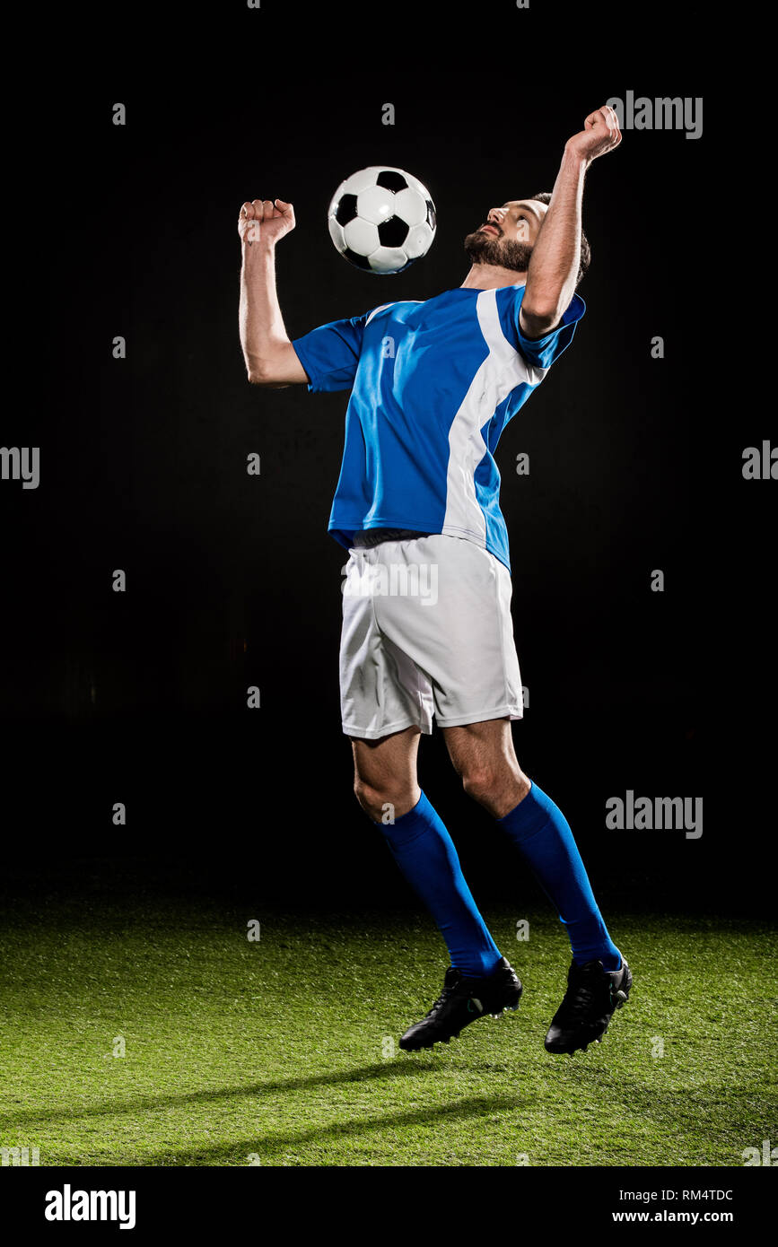 handsome football player in uniform jumping with ball isolated on black ...