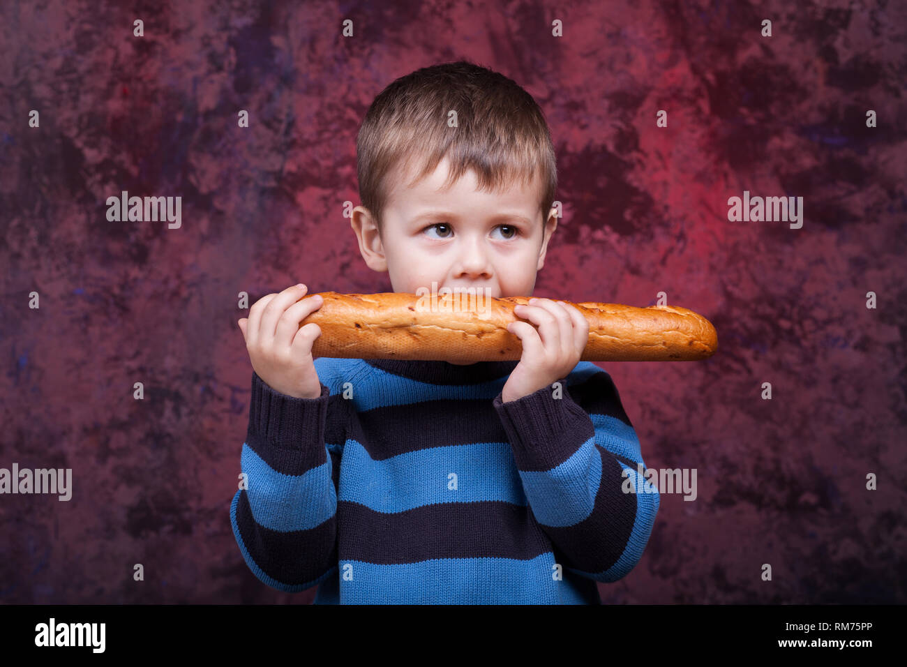 Cute kid holding and biting French bread against dark red background ...