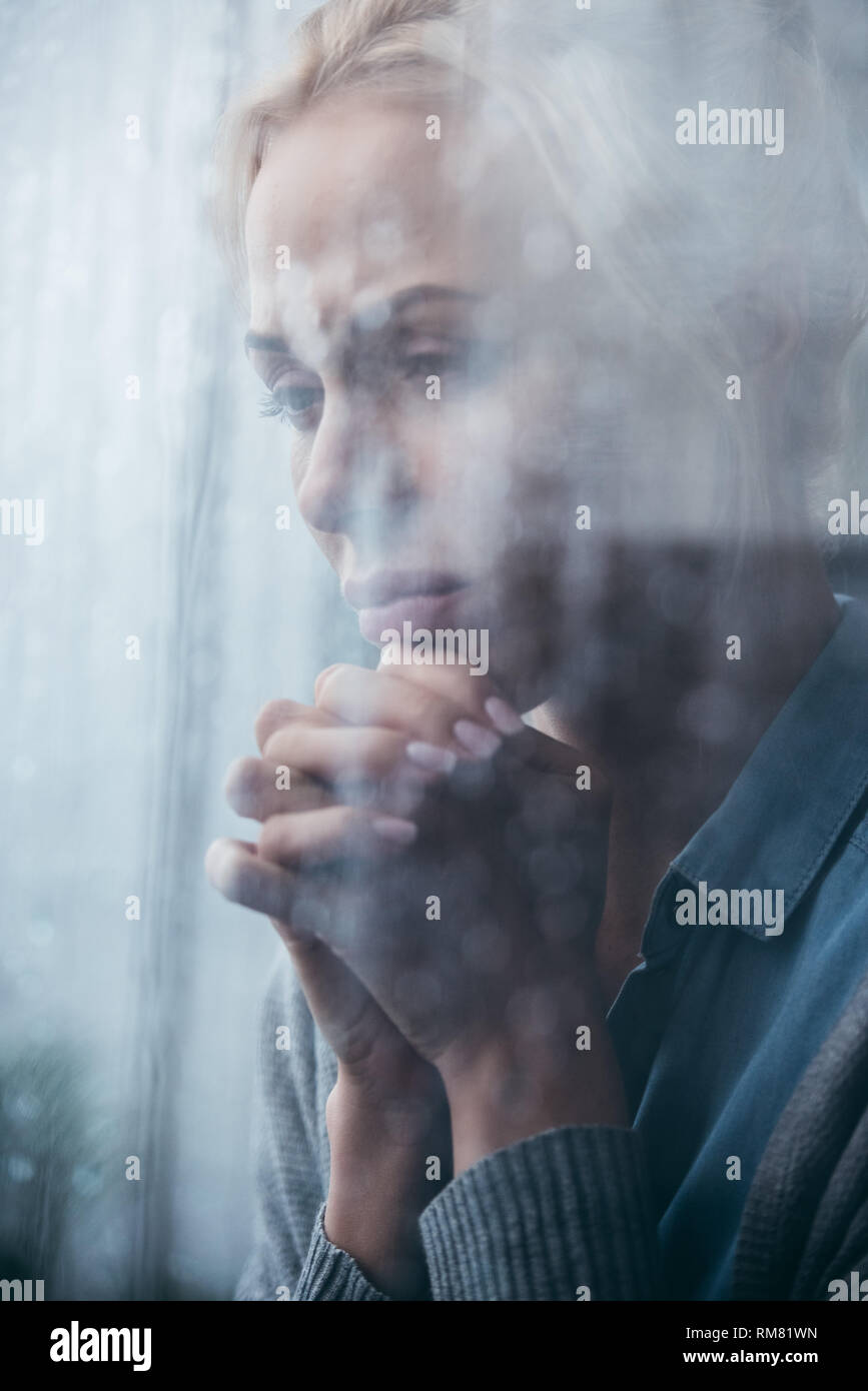 sad adult woman with folded hands at home through window with raindrops ...