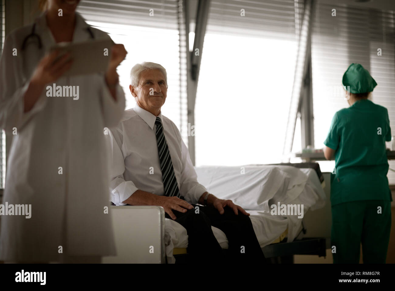 View of an elderly man sitting on a bed as two doctors stand by Stock ...