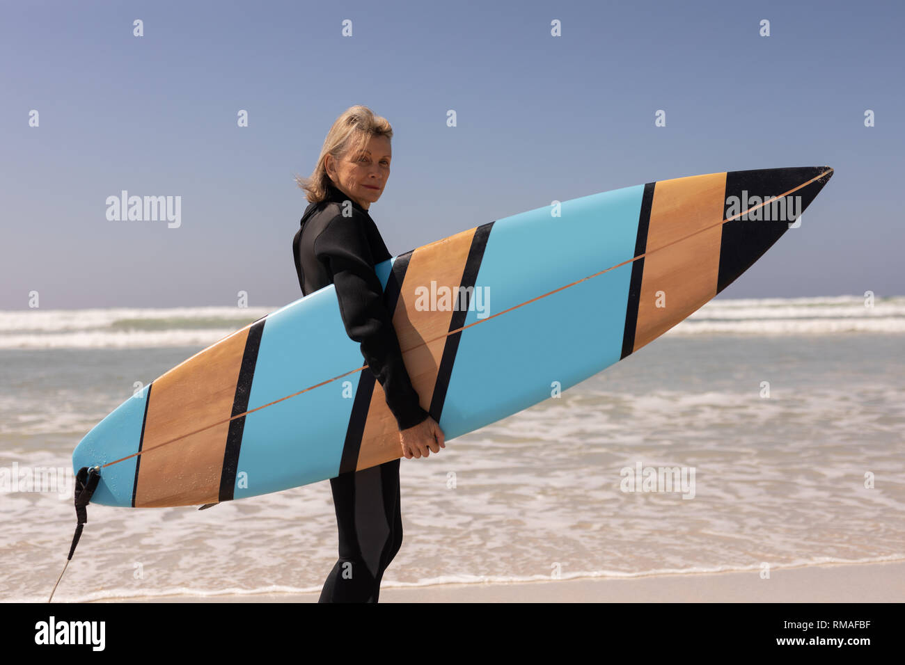 Side view of senior female surfer standing with surfboard on the beach ...