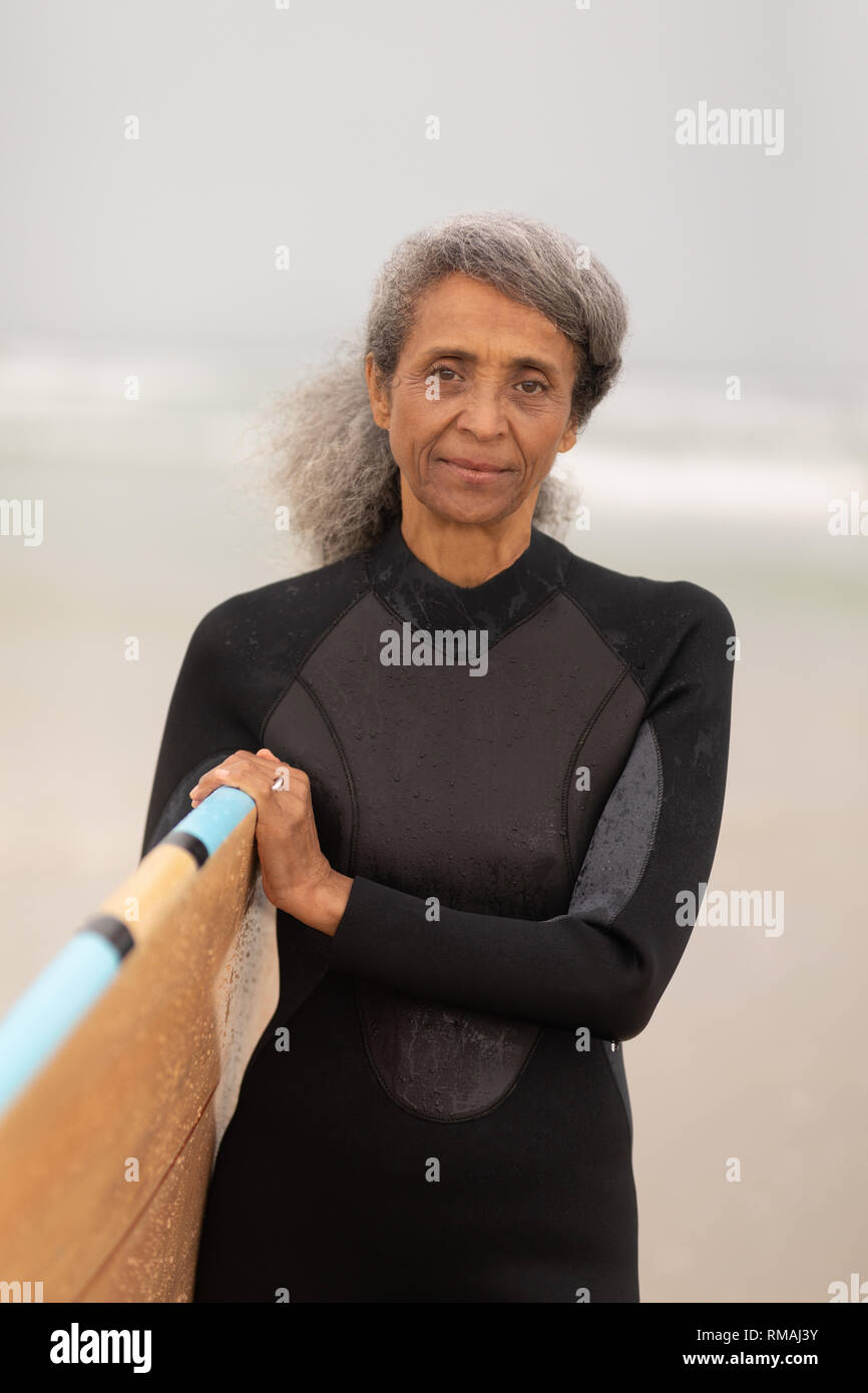 Senior female surfer standing with surfboard and looking at camera on ...