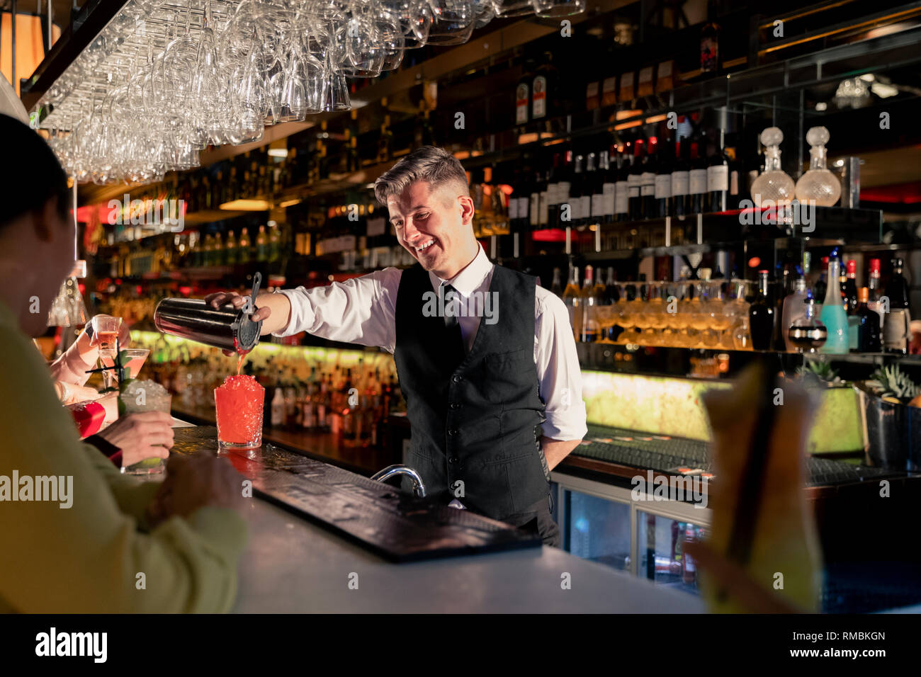 Young bartender pouring a cocktail out of a coktail shaker at the bar ...