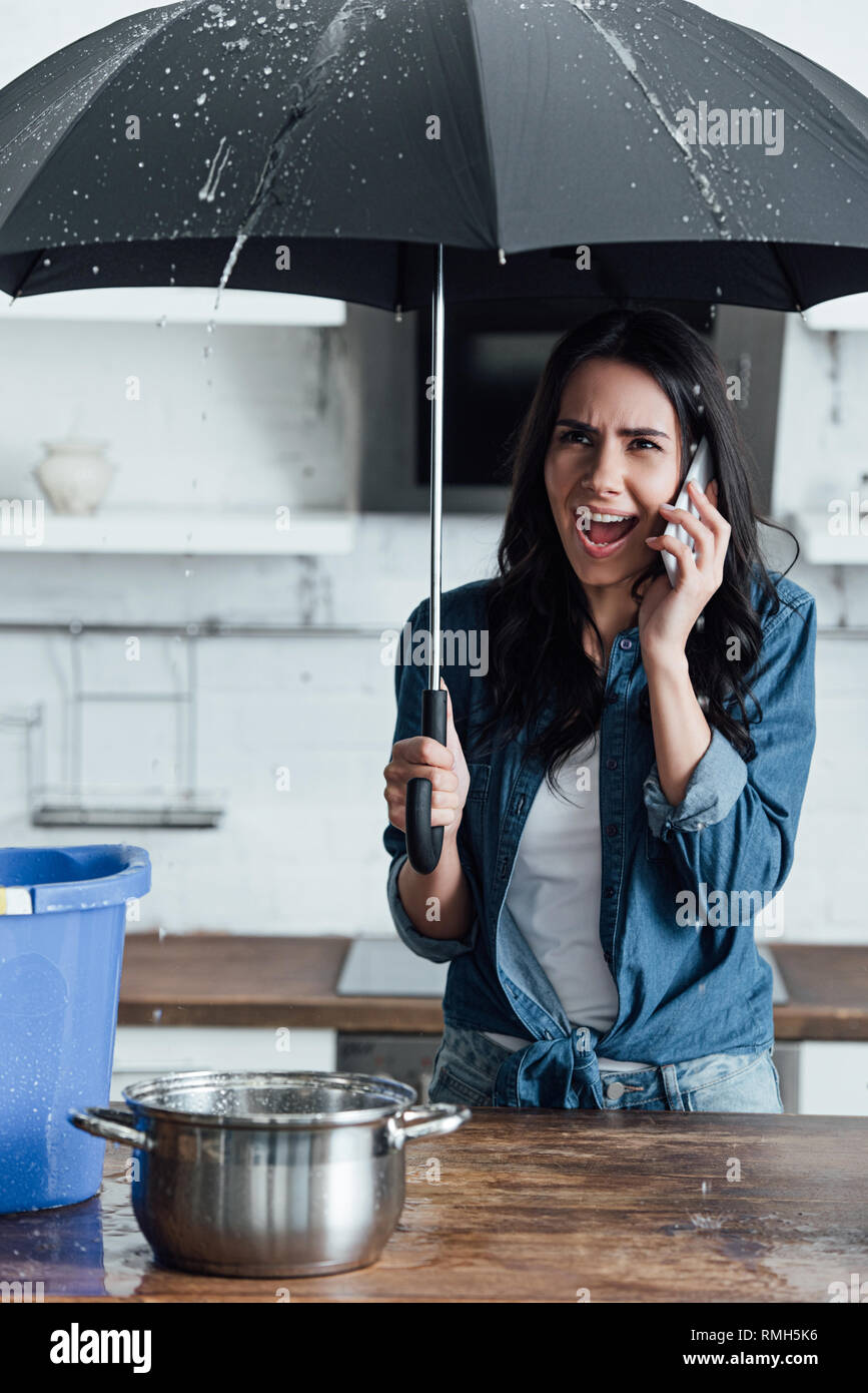 Stressed woman using umbrella during leak in kitchen and calling ...