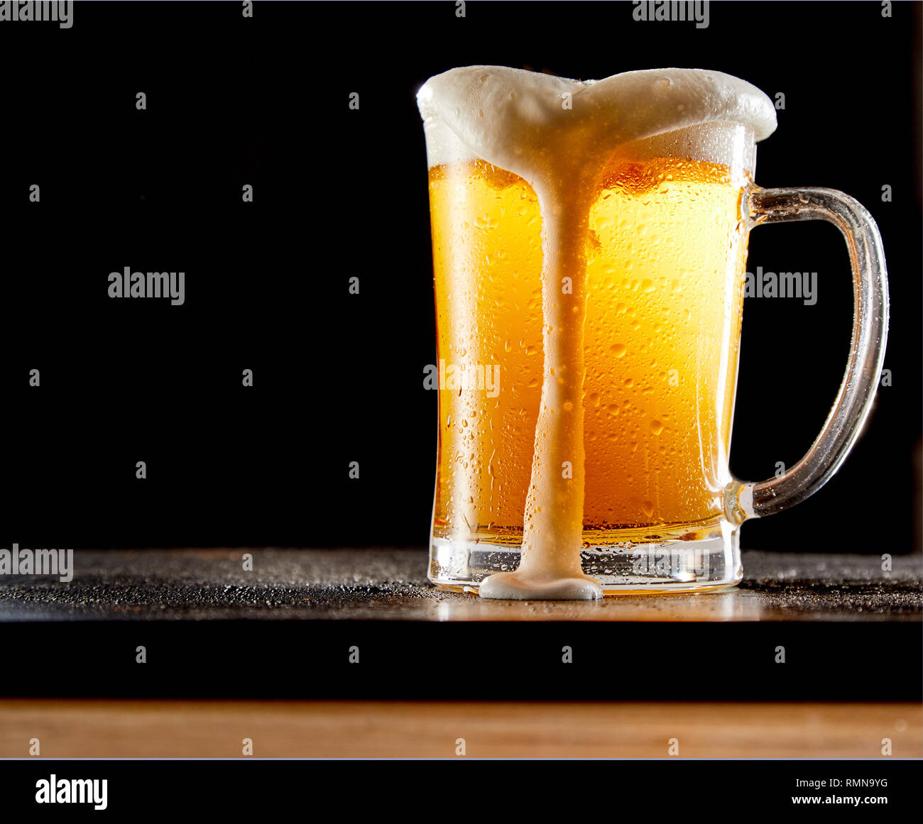 Mug of pale beer with dripping foam standing against black background ...