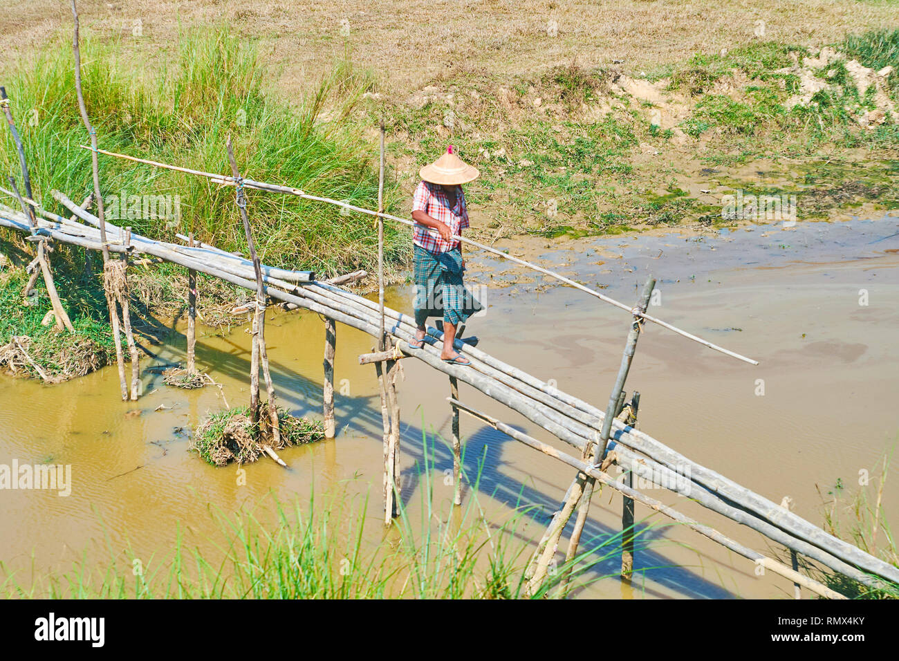 Burmese peasant in conical hat crosses primitive bamboo footbridge over ...