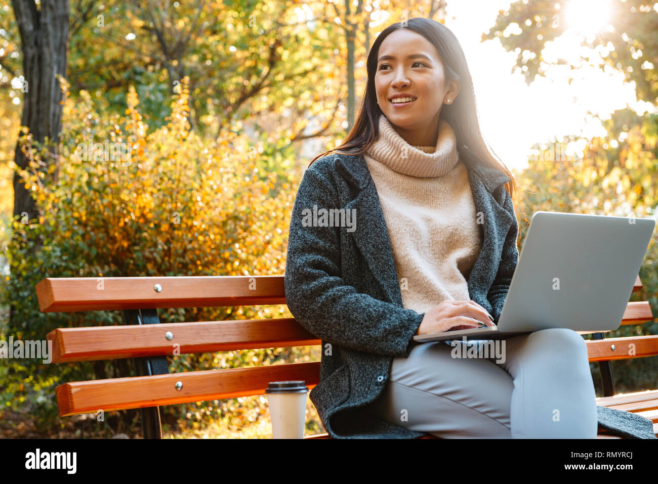 Smiling young asian woman wearing coat sitting on a bench at the park ...