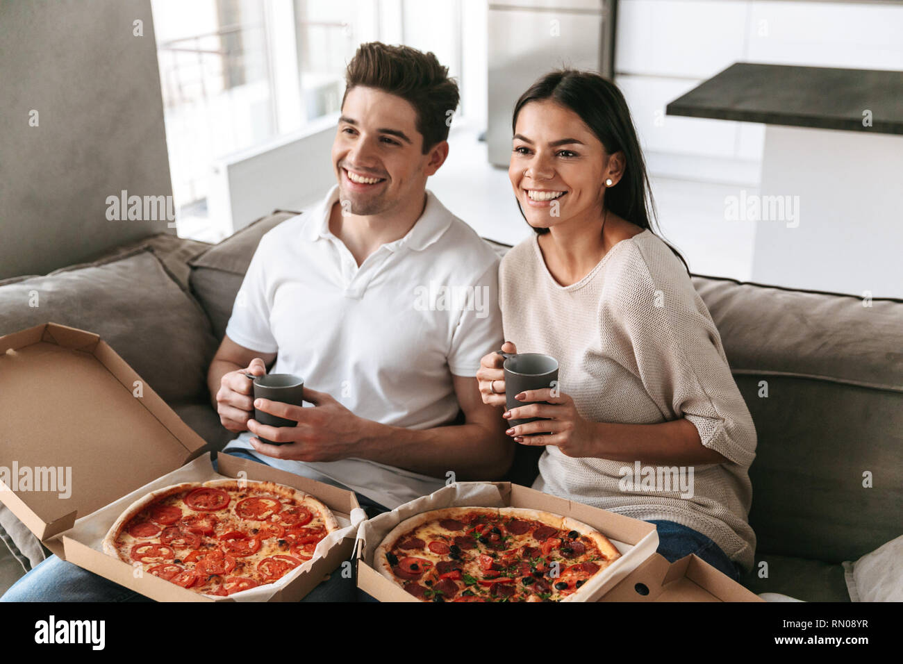Cheerful young couple sitting on a couch at home, eating pizza ...