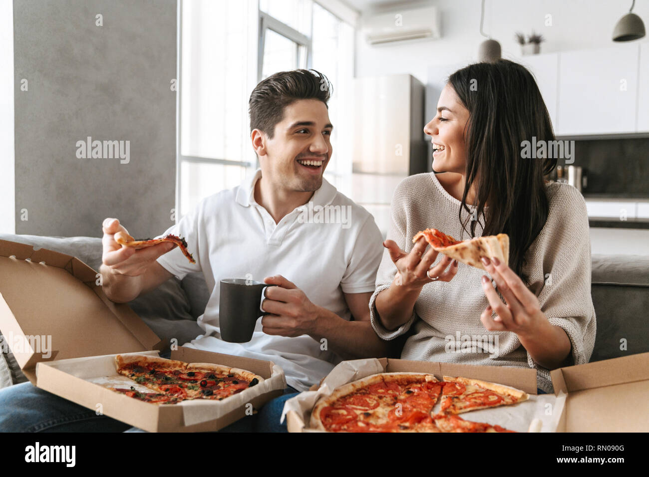Cheerful young couple sitting on a couch at home, eating pizza ...
