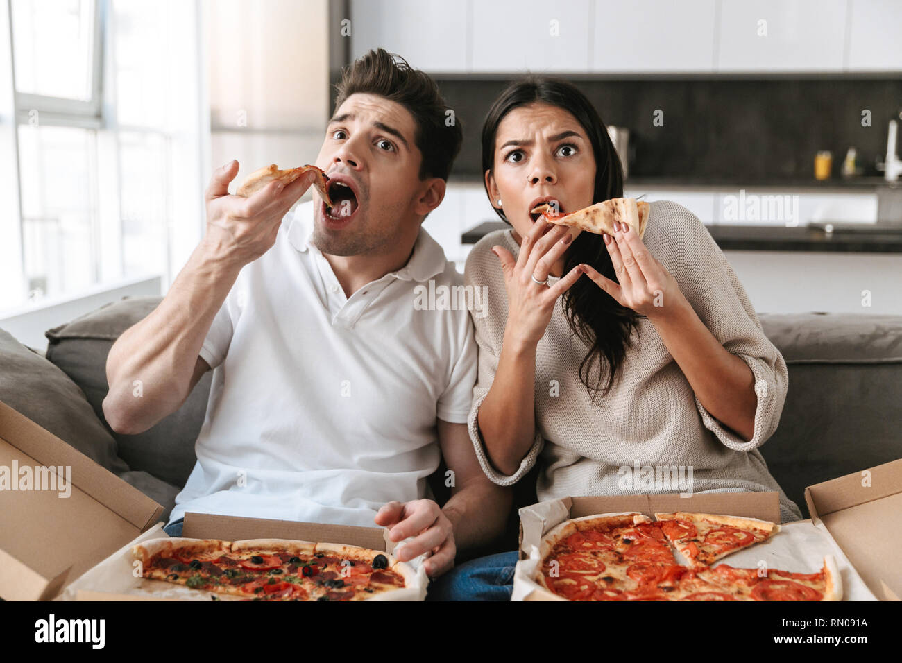 Cheerful young couple sitting on a couch at home, eating pizza ...