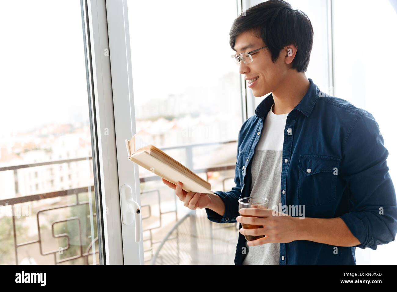 Smiling asian man reading a book while standing at the window at home ...