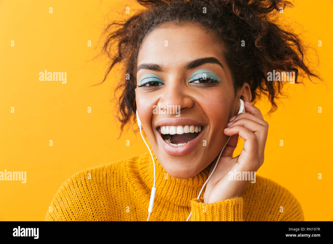 Cheerful african woman wearing sweater standing isolated over yellow background, listening to ...