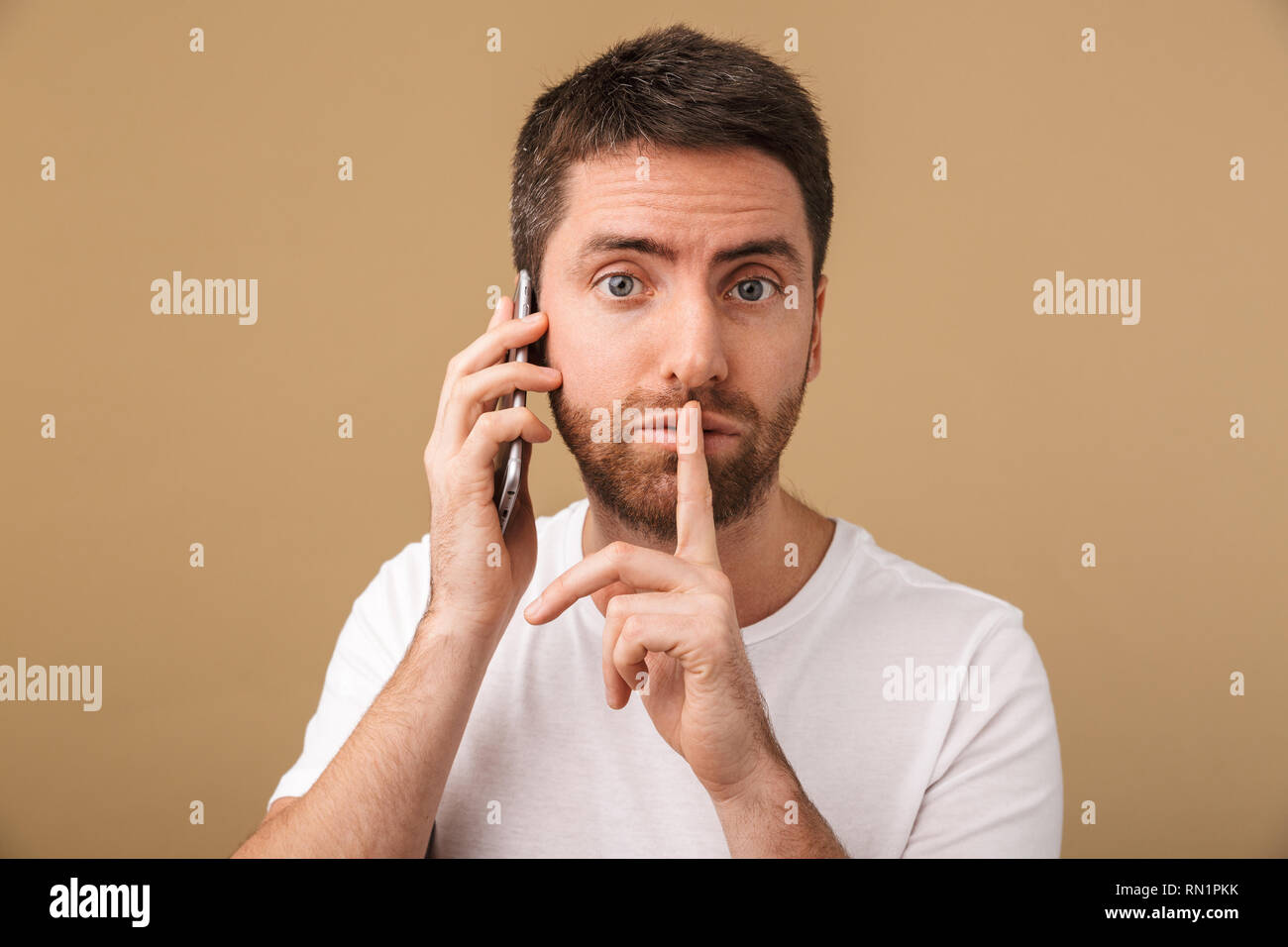Serious young casual man talking on mobile phone isolated over beige ...