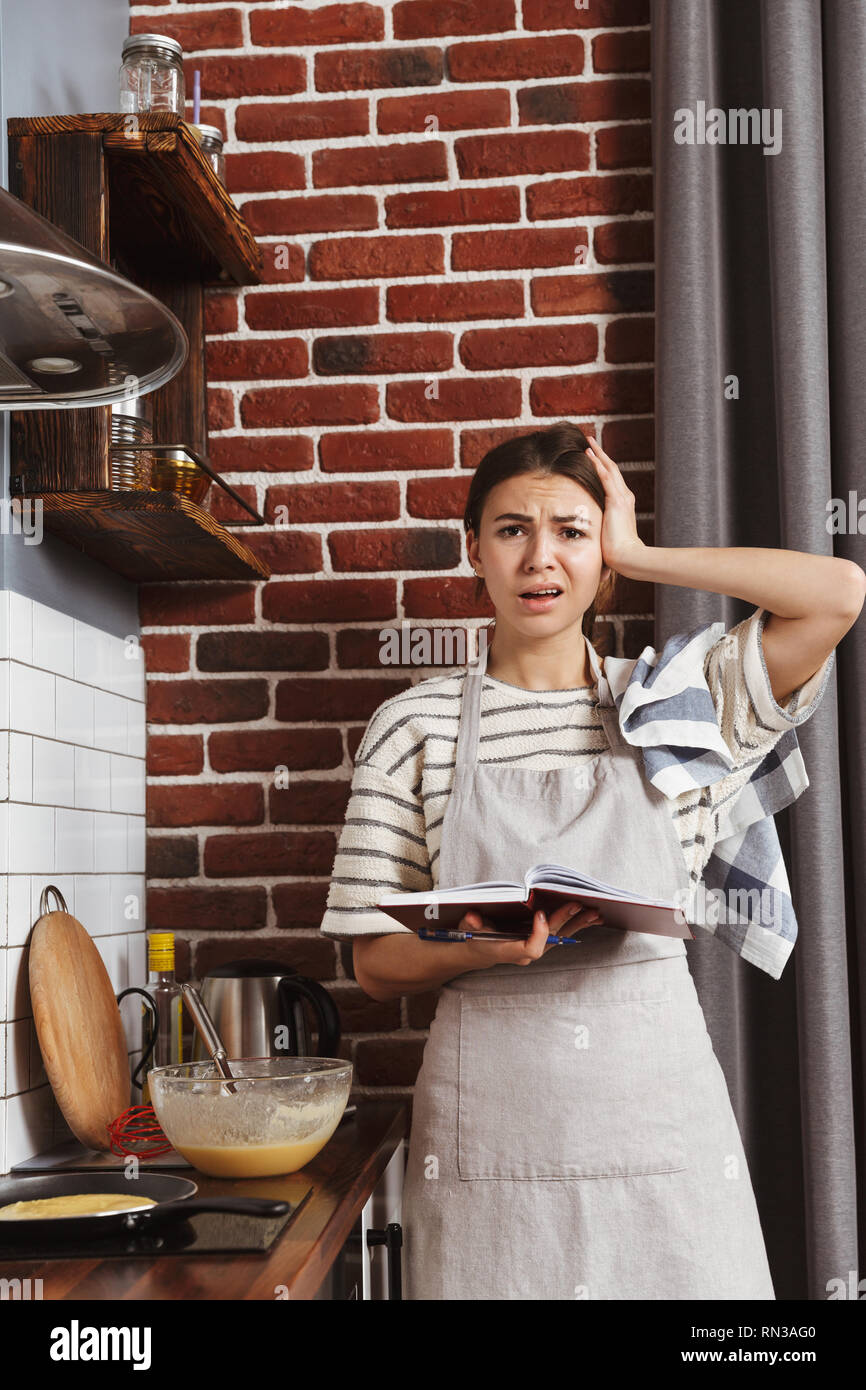 Confused young woman standing at the kitchen at home, cooking pancakes ...