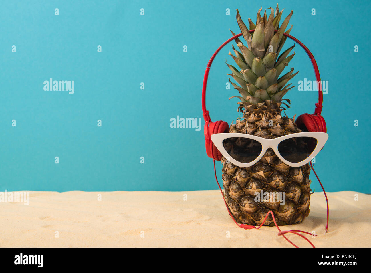 pineapple with headphones and sunglasses on sand on blue background ...