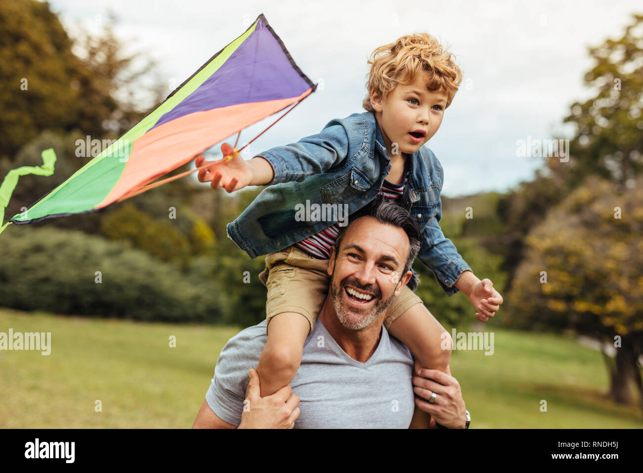 Little boy holding a kite sitting on his father's shoulders at the park ...